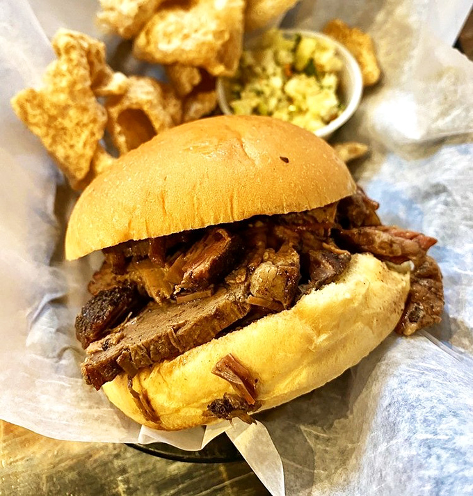 The pulled pork sandwich – barbecue in its most honest form. Tender strands of smoke-infused pork on a simple bun, with crispy skin cracklins that shatter like porcine candy.