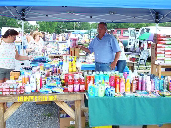 A vendor chats with customers at Leighty's Flea Market, where tables are stocked with personal care products, snacks, and bargain finds under a canopy on a sunny day.