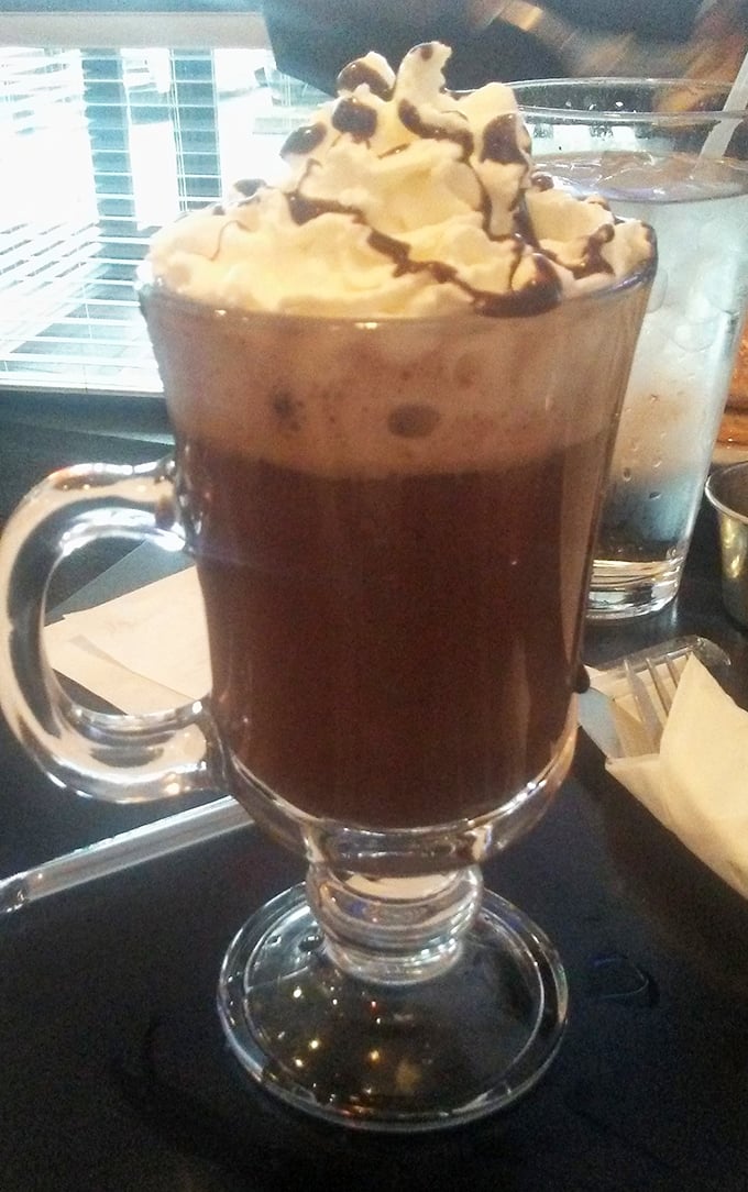 Hot chocolate that understands its assignment—topped with a cloud of whipped cream and chocolate drizzle in a glass tall enough to impress even your sweet tooth.