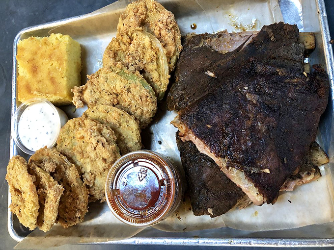 A tray of ribs with the perfect bark, flanked by golden cornbread and fried green tomatoes – the holy trinity of Southern comfort on a metal tray.