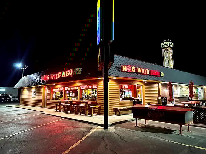 The red neon glow of Hog Wild BBQ beckons hungry travelers like a lighthouse for the barbecue-starved soul. The rustic log cabin exterior promises authentic, no-frills deliciousness within.