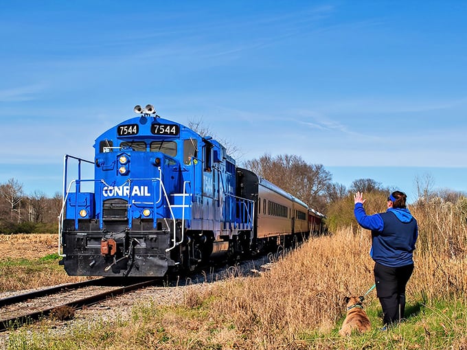 The blue Conrail locomotive powers through Ohio countryside as a spectator and furry friend wave hello – a perfect snapshot of railroad enthusiasm in action.