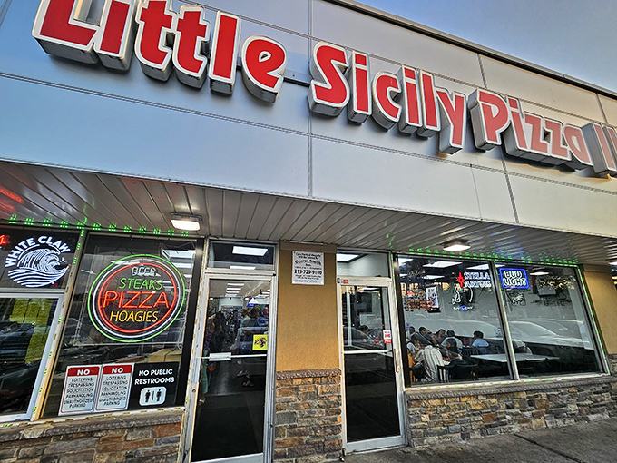 The daytime view reveals the restaurant's no-nonsense exterior, where that neon "STEAKS PIZZA HOAGIES" sign tells you everything you need to know.
