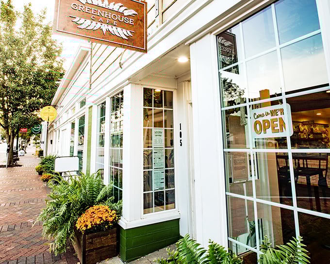 The Greenhouse Cafe's inviting storefront beckons with its crisp white exterior, wooden sign, and those vibrant orange mums that practically shout "Come in and stay awhile!"