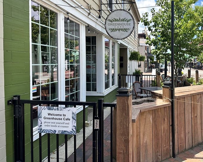 The Greenhouse Cafe welcomes you with its charming green exterior, classic white-framed windows, and a cozy outdoor seating area framed by natural wood—inviting you to come in, relax, and enjoy.