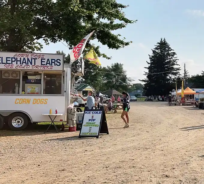 A casual country market scene where food trucks serve up corn dogs and elephant ears, setting the stage for a day of small-town treasure hunting and bargain finds under the Michigan sun.