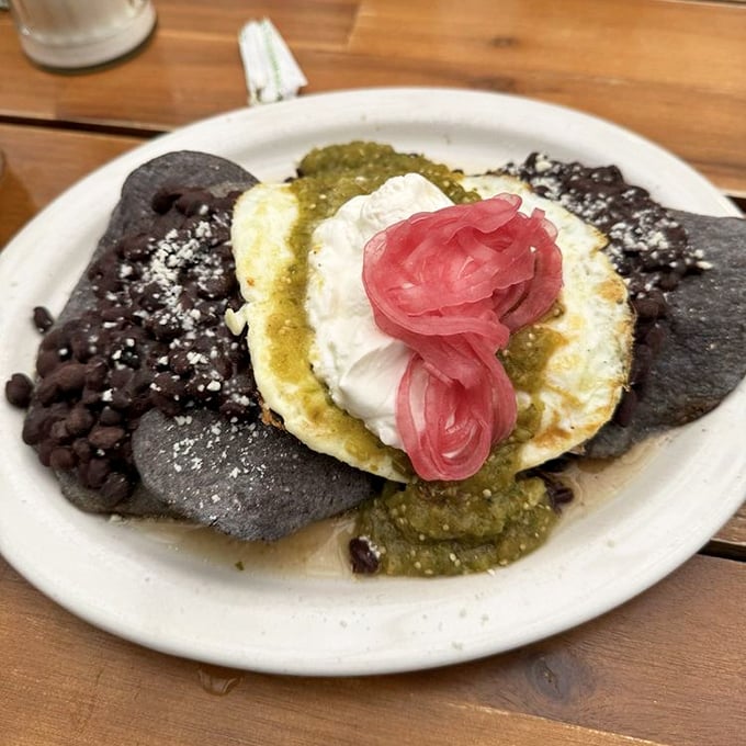 Black bean tortillas cradle a sunny-side egg crowned with salsa verde and pickled onions – breakfast reimagined with artistic flair.