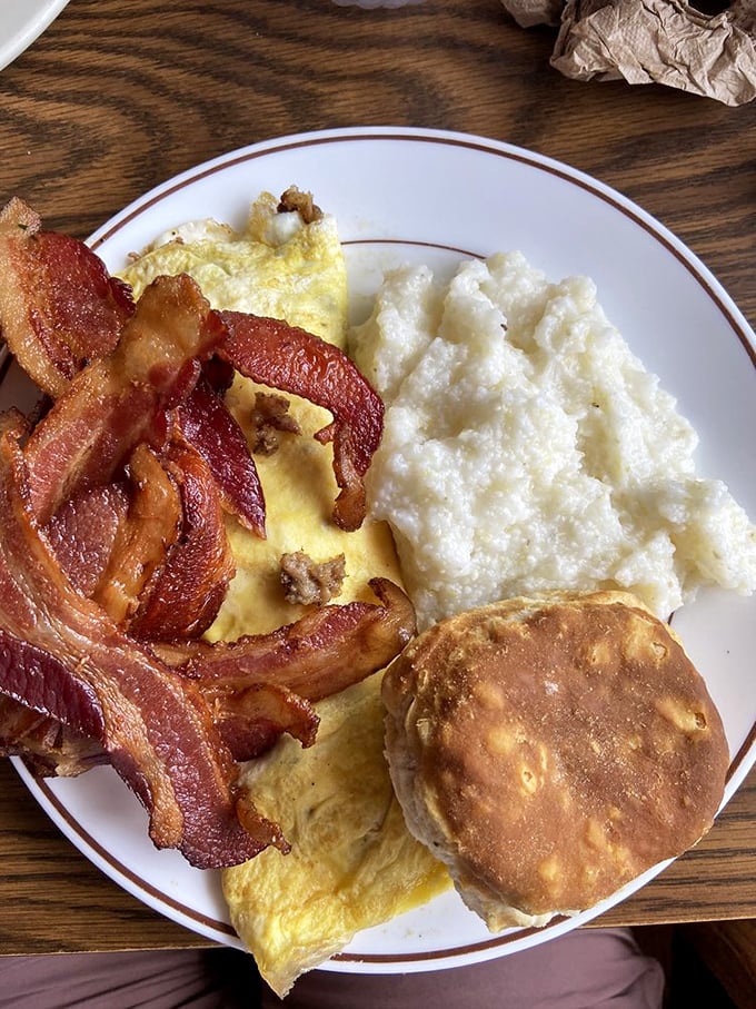 The breakfast trinity: crispy bacon, fluffy eggs, and grits with a golden biscuit standing guard. This plate doesn't need a prayer—it is the blessing.