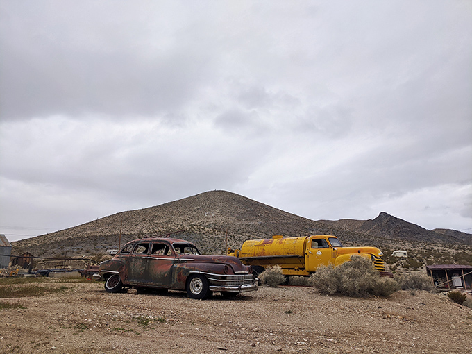 A vintage car and yellow truck stand sentinel against the mountain backdrop—desert sculptures with stories to tell.
