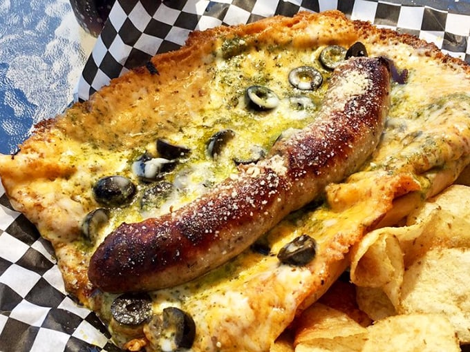 The Pesto Brat redefines sausage expectations – nestled in a golden cheese blanket with olives standing guard over this flavor masterpiece.