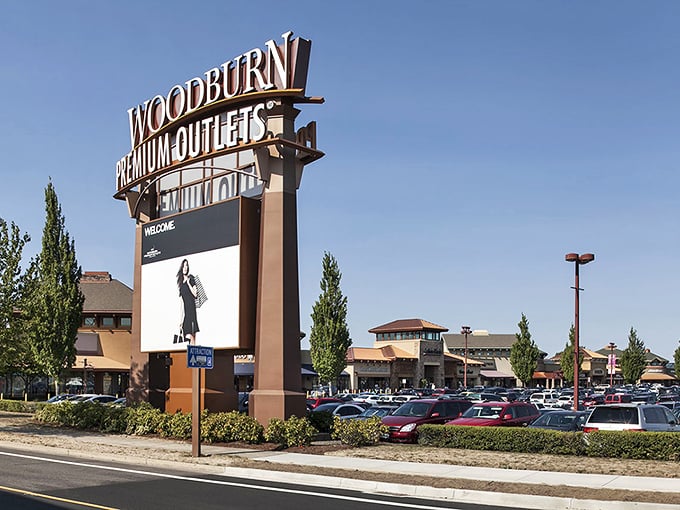 The expansive parking lot stretches toward retail horizons. Car trunks enter empty and leave full—the universal sign of a successful outlet adventure.