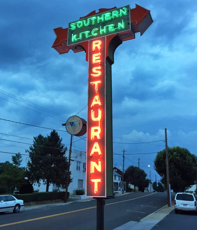As evening falls, the neon sign glows like a lighthouse for hungry travelers – "SOUTHERN KITCHEN RESTAURANT" in electric red and green against the twilight sky.