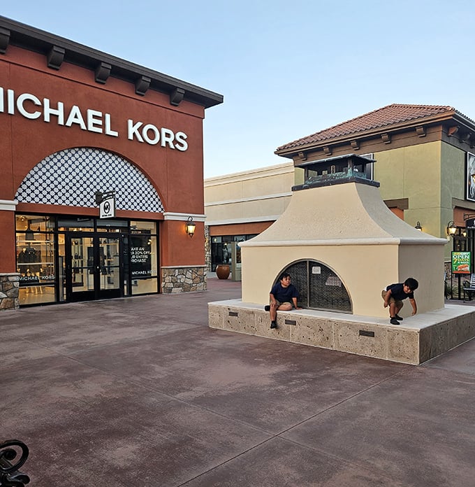 The Michael Kors storefront stands proudly in terra cotta splendor, while children discover the simple joy of a decorative fireplace structure—shopping and entertainment in one stop!
