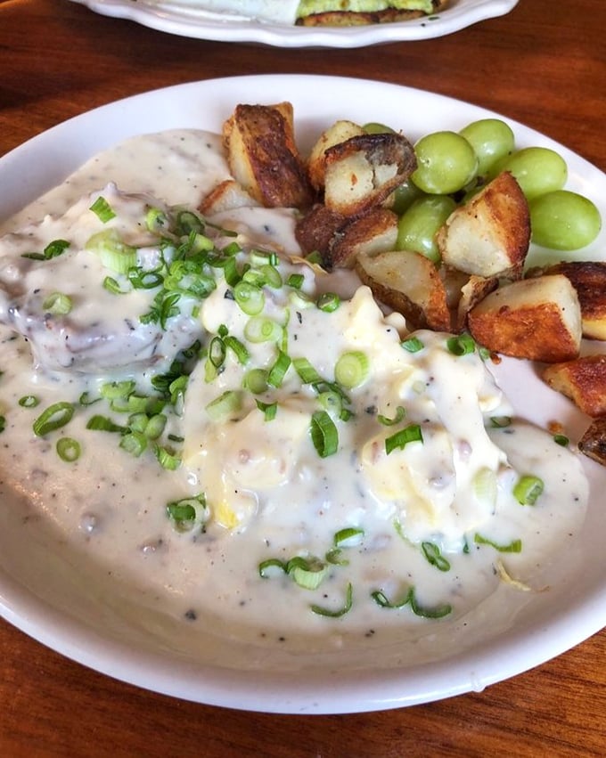 Behold the legendary biscuits and gravy—a velvety blanket of sausage gravy atop scrambled eggs, with crispy potatoes and grapes providing perfect counterpoints. 