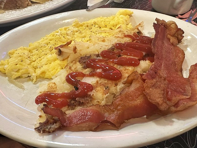 Breakfast architecture at its finest—scrambled eggs so fluffy they need anchoring, bacon with that perfect crisp-to-chew ratio, and ketchup applied with artistic precision.