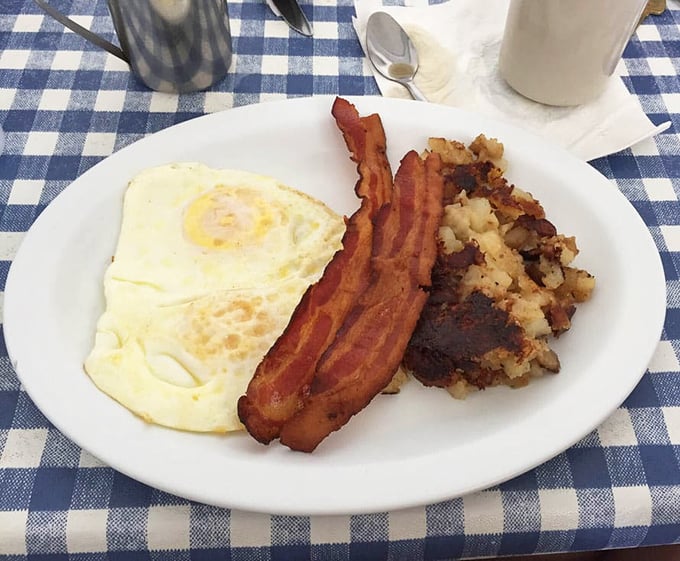 Breakfast perfection doesn't need fancy plating – just a sunny-side up egg, crispy bacon, and home fries on a blue-checked tablecloth.