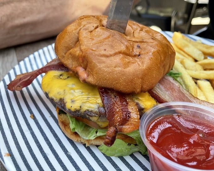 Behold the burger in its natural habitat – draped in melted cheese, adorned with bacon, and stabbed dramatically with a knife as if to say, "Challenge accepted?"