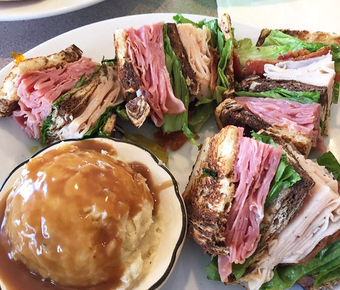 Behold the club sandwich in all its triple-decker glory! Layers of meat and freshness stacked between perfectly toasted bread, with gravy-smothered potatoes alongside.