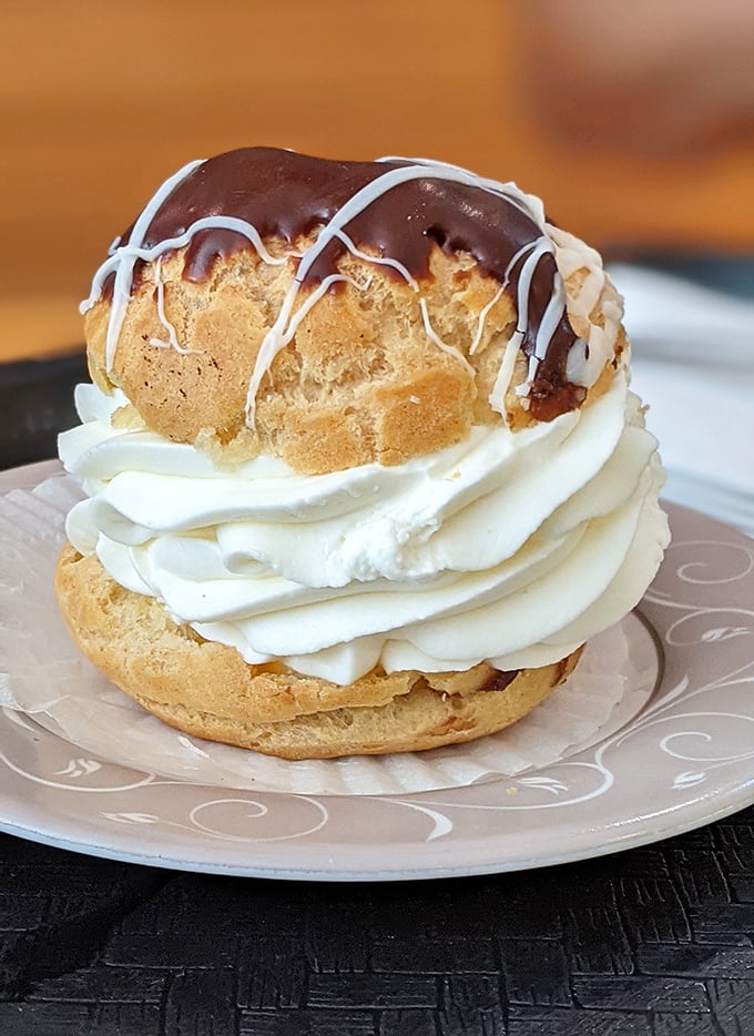 Behold the cream puff in its natural glory – a golden, chocolate-drizzled dome atop a cloud of whipped cream. Architecture this perfect deserves its own ZIP code.