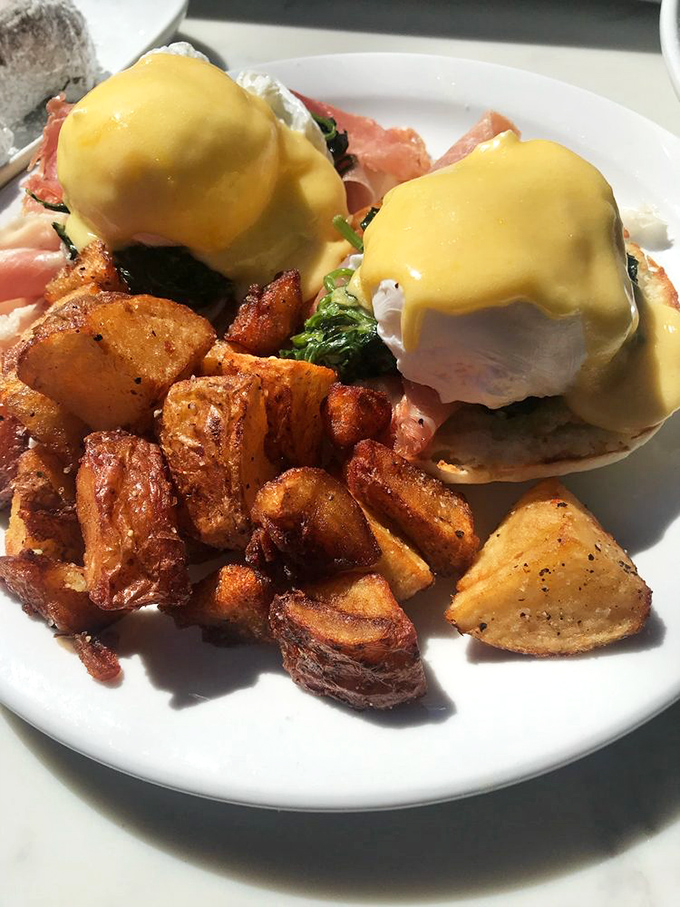 Eggs Benedict nirvana achieved: perfectly poached eggs draped in sunshine-yellow hollandaise, with crispy potatoes that demand equal attention.