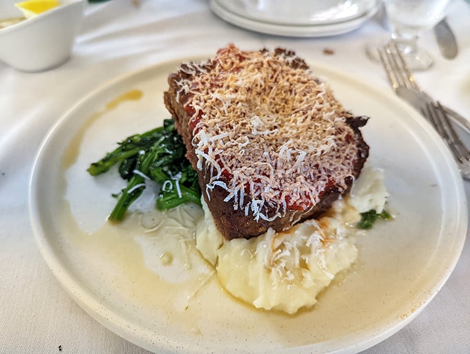 Not just meatloaf, but a masterpiece – crowned with cheese and nestled on cloud-like mashed potatoes with vibrant greens for balance.