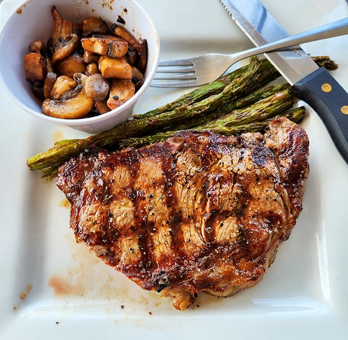 Behold the ribeye in its natural habitat – perfectly charred, resting beside grilled asparagus and sautéed mushrooms. This is meat's highest calling.