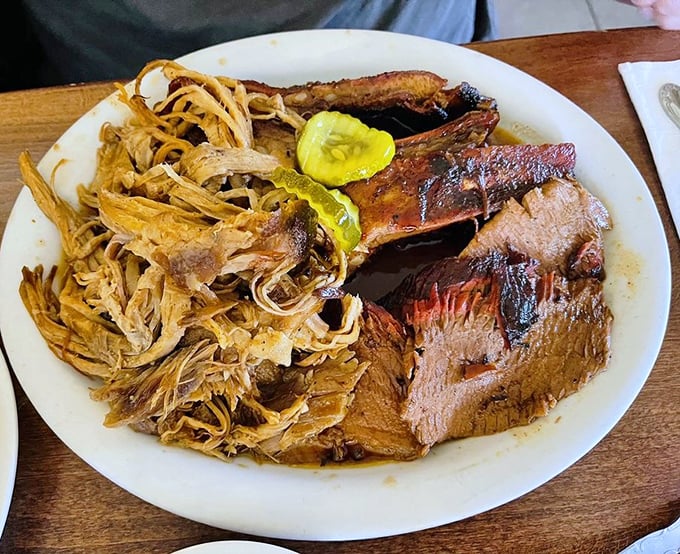 Behold the holy trinity of BBQ—brisket, pulled pork, and ribs sharing a plate in perfect harmony. Those pickle chips aren't just garnish; they're necessary acid relief.
