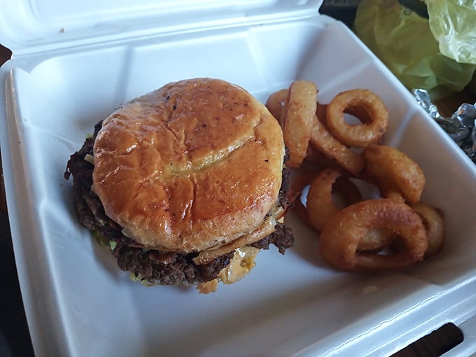 A classic diner burger nestled beside crispy onion rings in a takeout container – proof that simple food done right needs no fancy presentation.