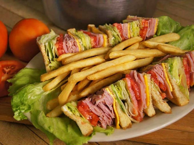 The club sandwich—that architectural marvel of deli meat, fresh vegetables, and toasted bread—stands tall with a fortress of golden fries as its loyal guards.