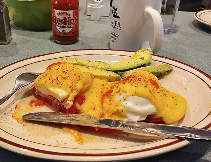Eggs Benedict with a side of avocado—hollandaise cascading down like a golden waterfall. Frank's RedHot nearby suggests some diners like their morning with a kick.