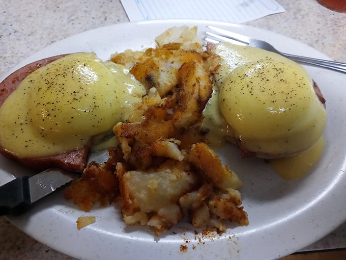 Eggs Benedict done right – perfectly poached eggs wearing hollandaise like a silk robe, perched atop Canadian bacon with crispy home fries standing guard.