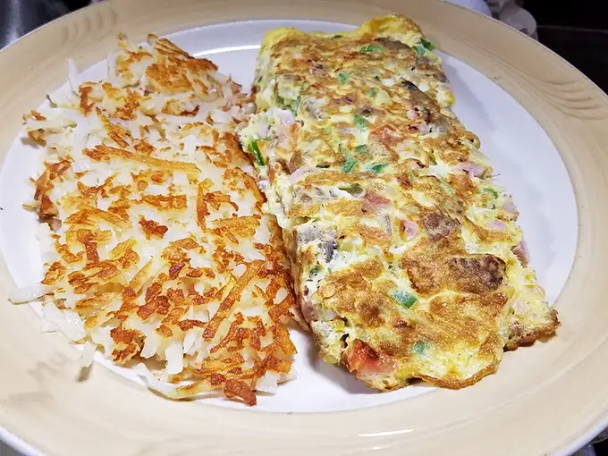 This omelet doesn't just sit on the plate—it commands attention with perfectly folded eggs alongside hash browns crisped to golden perfection.