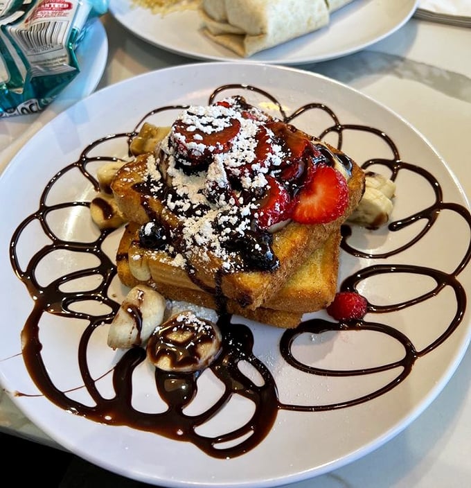 French toast elevated to art form—bananas, strawberries, chocolate drizzle, and powdered sugar create a breakfast that's basically dessert with permission.