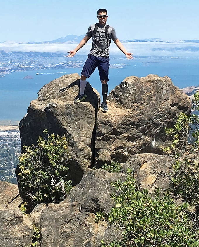 A hiker celebrates reaching a viewpoint that makes apartment security deposits seem reasonable again—fog-shrouded Bay Area splendor worth every step.