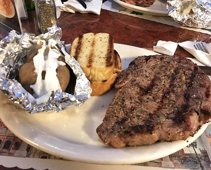 A plate that speaks the universal language of satisfaction – juicy steak, toasted garlic bread, and a foil-wrapped potato waiting to be unwrapped like a present.