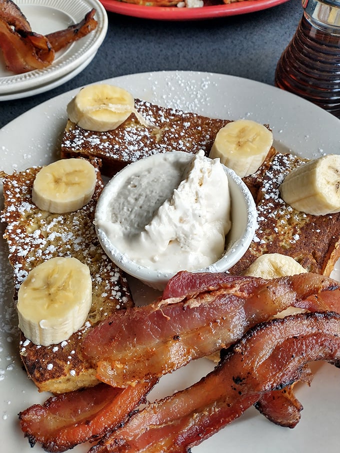 French toast that makes you question all other breakfasts – challah bread transformed into a banana-topped masterpiece with bacon standing guard.