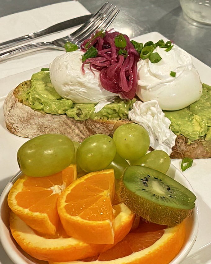 Avocado toast elevated from Instagram cliché to culinary masterpiece. Creamy avocado, perfectly poached eggs, and vibrant pickled onions atop artisanal bread with fresh fruit companions.