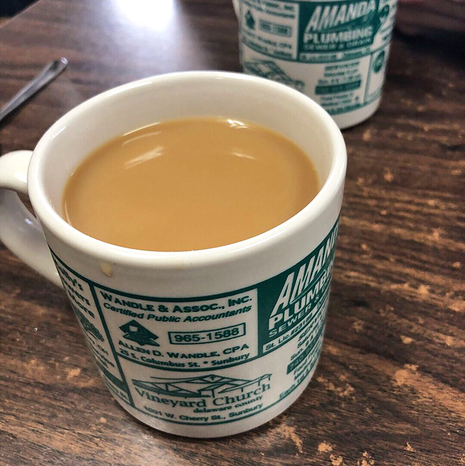The coffee comes in mugs advertising local businesses—a small-town tradition more authentic than any artisanal coffee shop's carefully curated aesthetic.