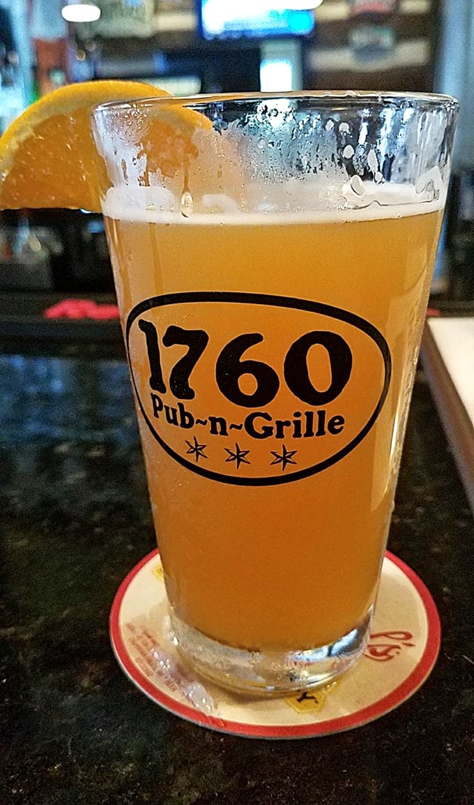 A perfect pour of golden craft beer, adorned with an orange slice, sits in a branded glass – liquid sunshine waiting to brighten your afternoon.