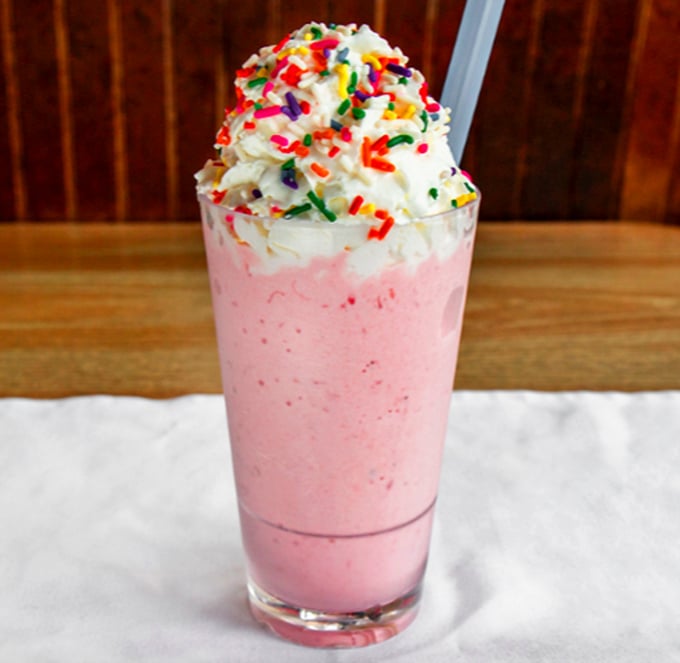 A strawberry milkshake topped with whipped cream and rainbow sprinkles—childhood nostalgia served in a glass tall enough for grown-up appetites.