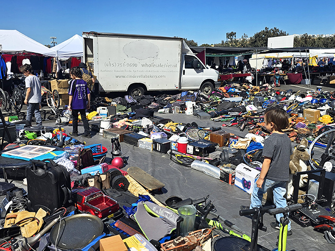 A bustling flea market filled with secondhand treasures—electronics, toys, tools, and clothes—sprawled out on tarps as families and bargain hunters browse under a bright blue sky.
