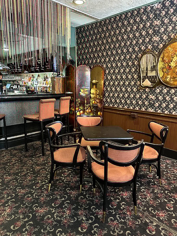 The bar area feels like stepping into a 1960s film set – patterned wallpaper, vintage dividers, and coral chairs creating a retro cocktail oasis.