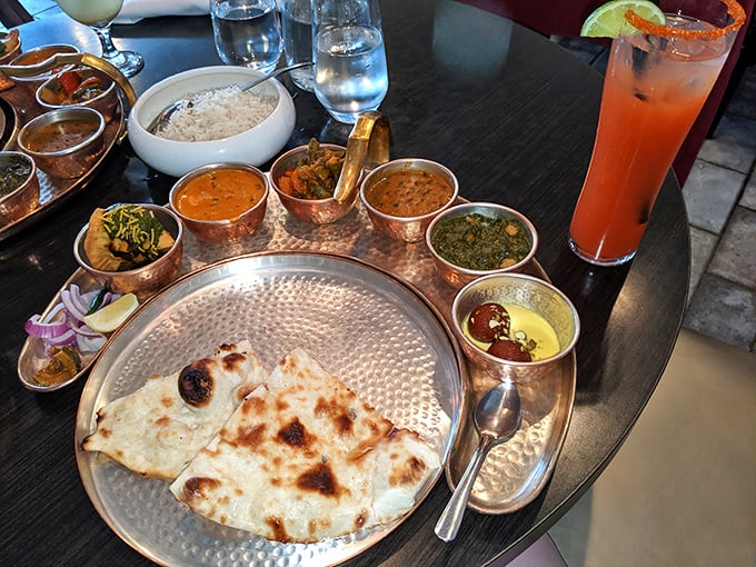 A copper-clad feast fit for royalty—curries in their burnished vessels surrounding freshly baked naan on a hammered silver platter, with a sunset-colored cocktail standing guard.