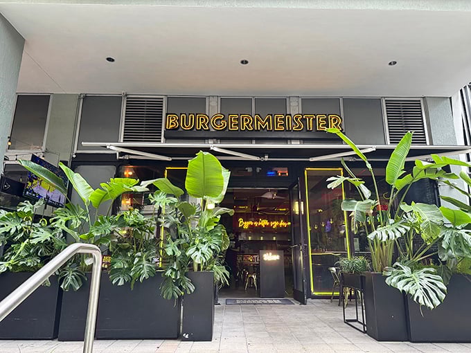 Burgermeister's entrance beckons with lush tropical plants and glowing yellow signage – like nature and neon joined forces to guide hungry humans home.