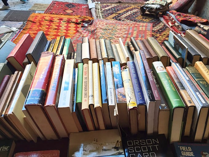 Used books stacked like literary lasagna, with vintage rugs as the backdrop. Somewhere in this pile is the novel that changes everything.