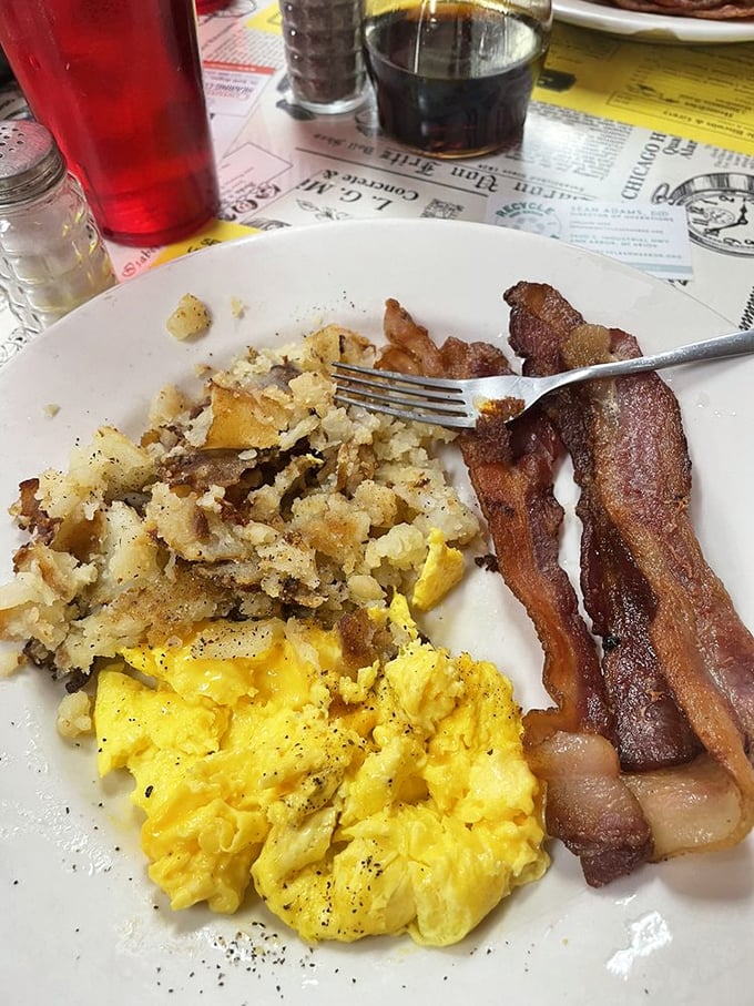 The breakfast trinity – golden eggs, crispy hash browns, and bacon cooked to that perfect point between chewy and crisp. Morning salvation on a white plate.