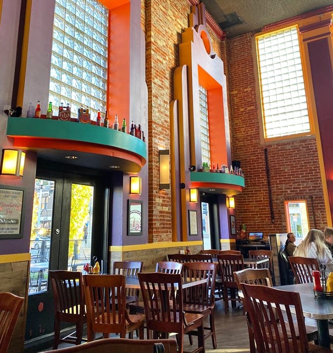 Sunlight streams through tall windows illuminating the retro-modern interior where brick, wood, and primary colors create the perfect backdrop for comfort food indulgence.