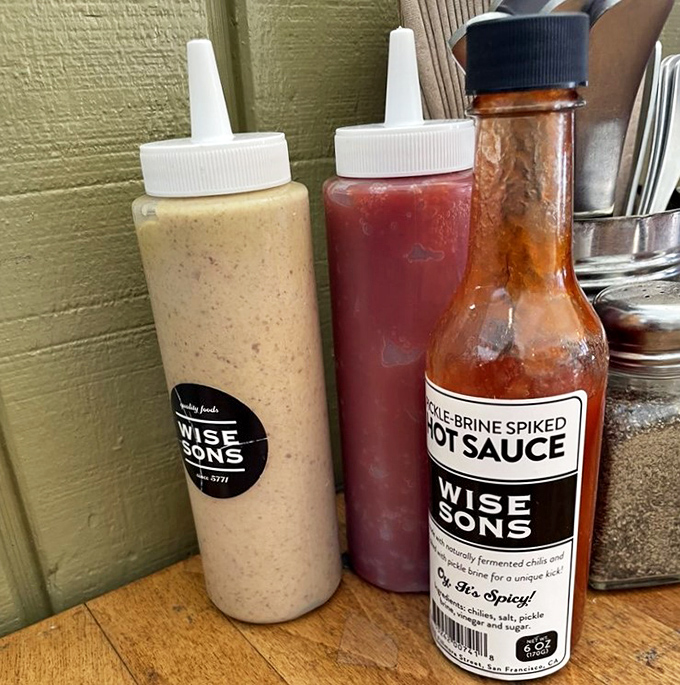 Condiment bottles standing at attention like delicious soldiers. The house-made pickle brine hot sauce adds a kick that would make your bubbe raise an eyebrow—in approval.