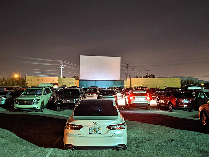 A sea of vehicles faces the blank canvas, headlights dimmed, everyone equal in the democracy of drive-in movie watching.