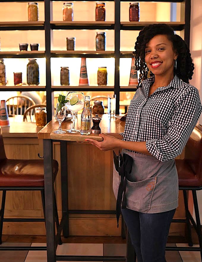 The display of preserved ingredients behind her isn't just decoration – it's a preview of the flavor complexity awaiting your palate.